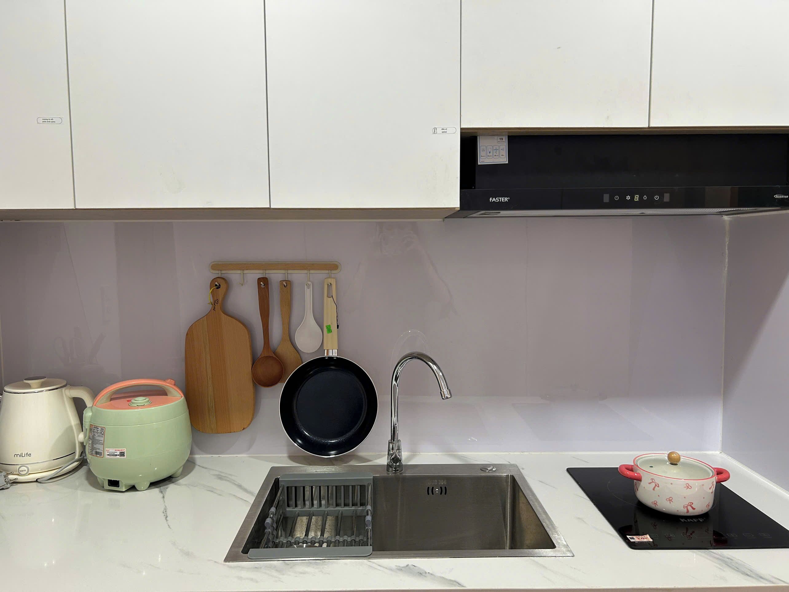 Uma cozinha com pia de aço inoxidável, chaleira, cozedor de arroz e panela em um fogão por indução.