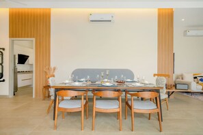 Uma sala de jantar com uma mesa posta para quatro pessoas, rodeada por cadeiras de madeira com almofadas cinzentas.