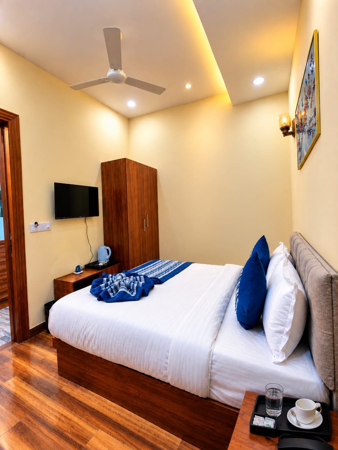 Quarto de hotel com cama, televisão, guarda-roupa, ventoinha no teto e um quadro na parede.