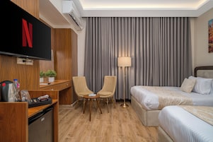 Quarto de hotel com televisão de tela plana, escrivaninha com cadeira, cama, luminária de chão e uma grande janela com cortinas.