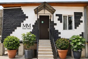 Entrada de hotel com design moderno, fachada em pedra, escada que leva à porta e plantas em vasos na entrada.