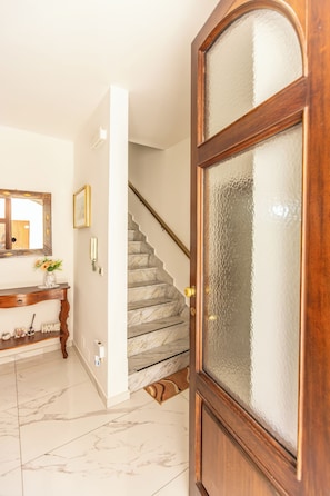 Uma escadaria com degraus de mármore que levam a um patamar, uma porta de madeira com painel de vidro jateado, um console com um vaso e um telefone fixado na parede.