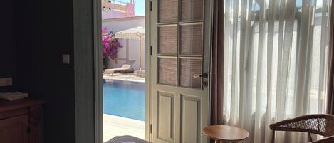Quarto de hotel com uma cama, uma cadeira, uma mesinha e uma porta para a área da piscina.