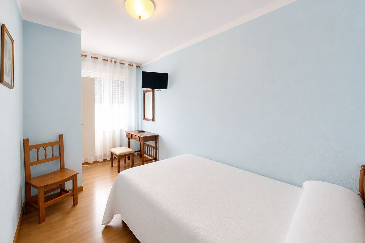 Quarto com cama, escrivaninha, cadeira e televisão na parede.