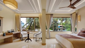 Quarto de hotel com cama, escrivaninha, cadeira, mesa e vista para palmeiras e edifícios pela janela.