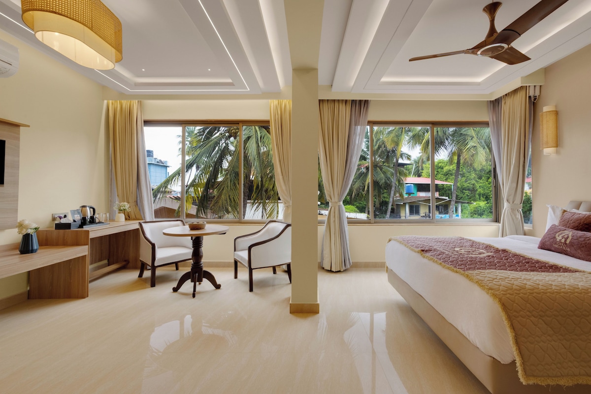 Quarto de hotel com cama, escrivaninha, cadeira, mesa e vista para palmeiras e edifícios pela janela.