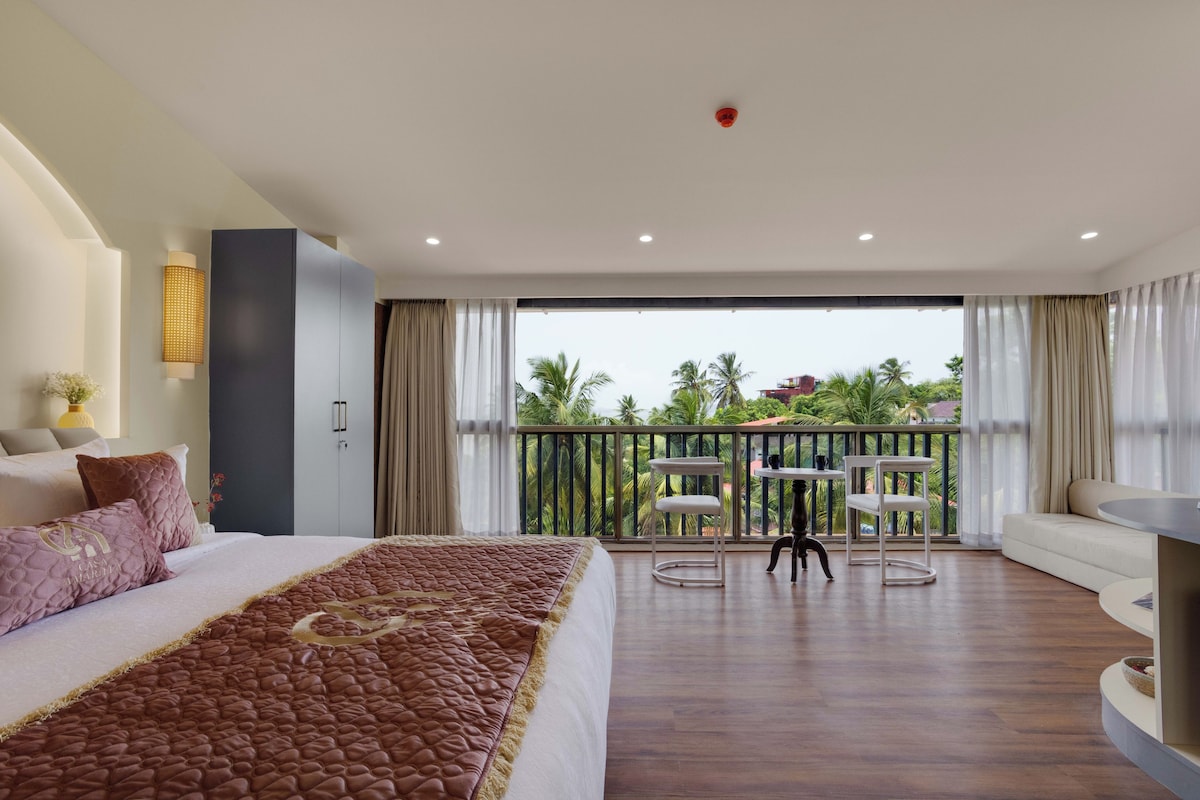Quarto de hotel com uma cama grande, varanda com mesa e cadeiras, e vista para palmeiras.