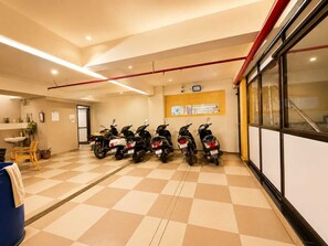 Um espaço interior limpo e bem iluminado, com chão quadriculado, uma fileira de scooters estacionadas, uma pia e uma janela com vista.