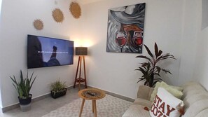 Uma sala de estar com televisão de tela plana, um quadro, uma luminária, uma mesa pequena e um sofá com almofada.