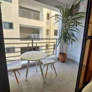 Uma varanda com mesa redonda e cadeiras, um vaso de planta e vista para um edifício com varandas.