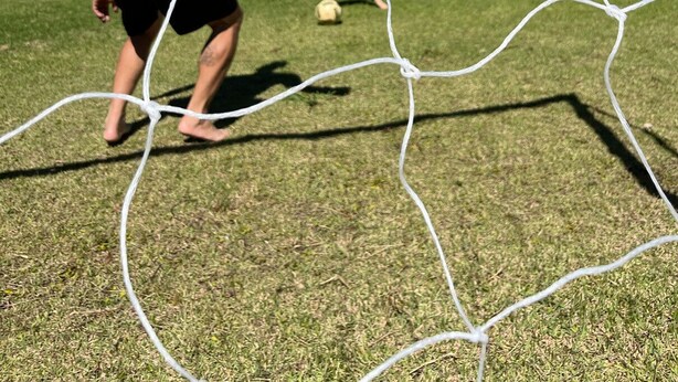 Jogo de futebol em andamento em campo gramado, com uma rede em primeiro plano.