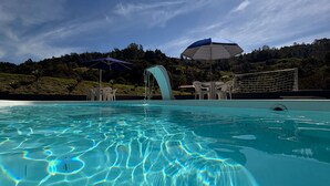 Piscina transparente com vista para uma colina e um edifício ao fundo.