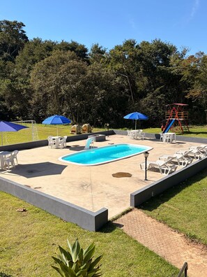 Piscina com escorregador, rodeada por cadeiras de lounge brancas e guarda-sóis, com árvores ao fundo.