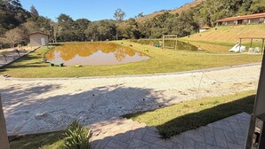 Vista de dentro de um edifício para uma área gramada com um lago, um pequeno galpão e uma colina com árvores ao fundo.