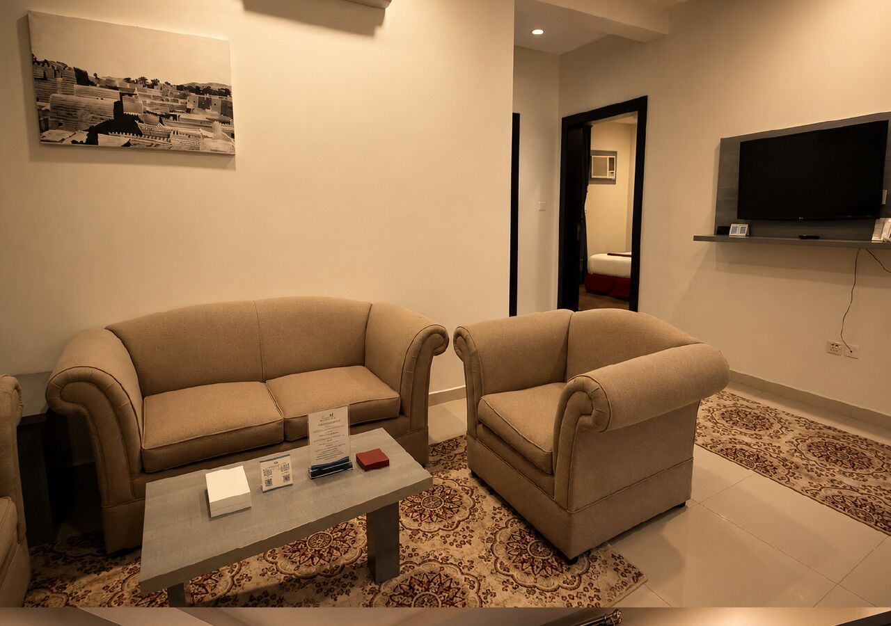Uma sala de estar com um sofá bege, uma mesa de centro com objetos, um tapete estampado e uma televisão de tela plana fixada na parede.