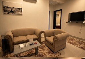 Uma sala de estar com um sofá bege, uma mesa de centro com objetos, um tapete estampado e uma televisão de tela plana fixada na parede.