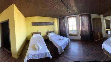 Quarto de hotel com duas camas de solteiro, uma cama de casal, um guarda-roupa, uma televisão e uma janela com cortinas.