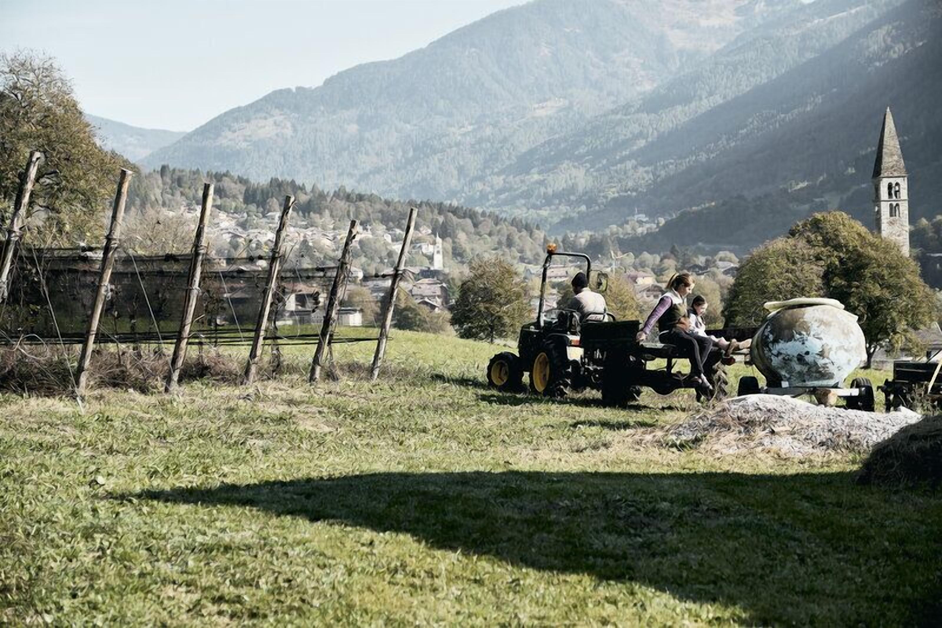 Paisagem rural com um trator, pessoas trabalhando em um campo, uma torre de igreja e montanhas ao fundo.