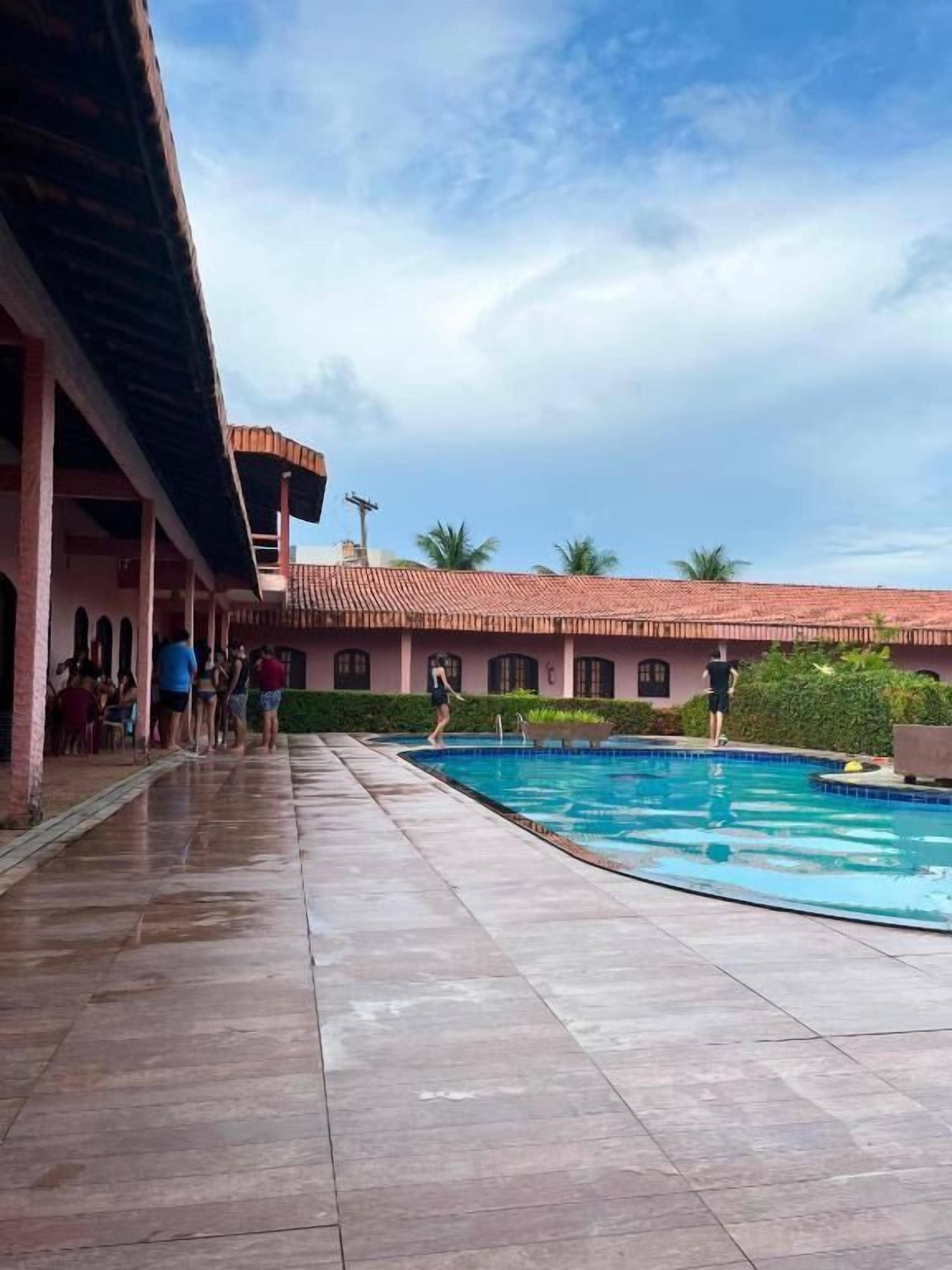 Área da piscina com uma galeria coberta e um edifício com janelas em arco ao fundo.