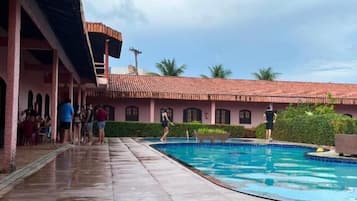 Área da piscina com uma galeria coberta e um edifício com janelas em arco ao fundo.