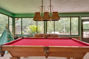 Uma mesa de bilhar com superfície de feltro vermelho, cercada por paredes verdes e grandes janelas.