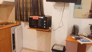 A compact room with a kitchenette, a television mounted on the wall, a small trash bin, and a desk with a chair.