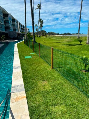 Piscina ao lado de um edifício, com área gramada e palmeiras.