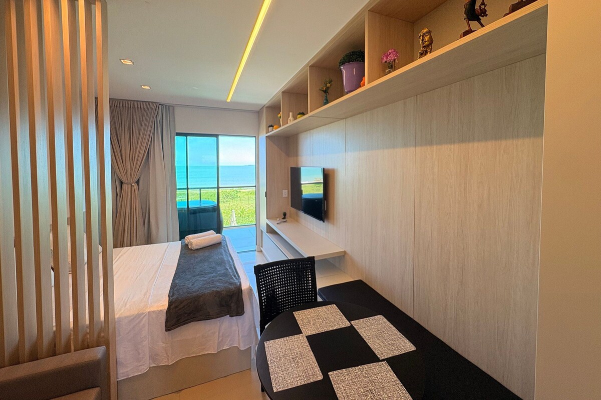 Quarto de hotel moderno com cama, um espaço de estar pequeno, televisão e vista para o mar.