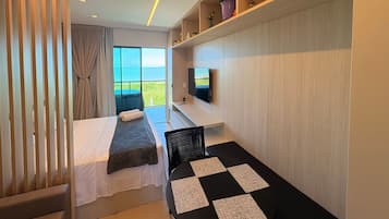 Quarto de hotel moderno com cama, um espaço de estar pequeno, televisão e vista para o mar.