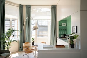 Uma sala de estar moderna com área de jantar, janelas amplas com cortinas, um abajur e uma cozinha com paredes verdes e uma pia.