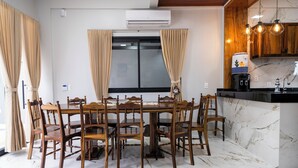 Uma área de refeições com mesa e cadeiras de madeira, um aparelho de ar condicionado acima da janela, e uma cozinha com piso de mármore e armários de madeira.