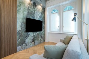Uma sala de estar moderna com uma televisão de tela plana fixada numa parede de mármore, piso de madeira, sofá cinza com almofadas e um abajur de chão.