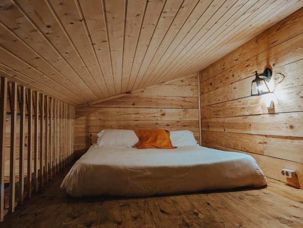 Quarto de cabana de madeira com uma cama, uma mesinha de cabeceira e uma luminária fixada na parede.