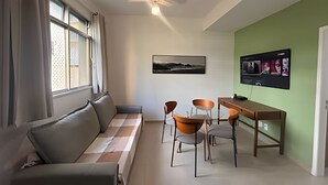 Uma sala de estar moderna com sofá, mesa de jantar e cadeiras. Há um ventilador de teto, uma janela com cortinas e uma televisão na parede.