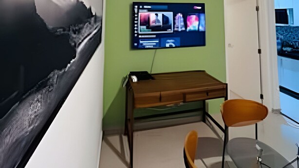 Quarto com televisão na parede, uma escrivaninha e uma cadeira.