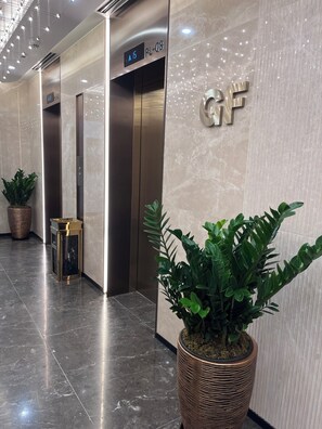 Área de elevadores moderna, com painel digital, balcão de recepção e plantas em vasos.