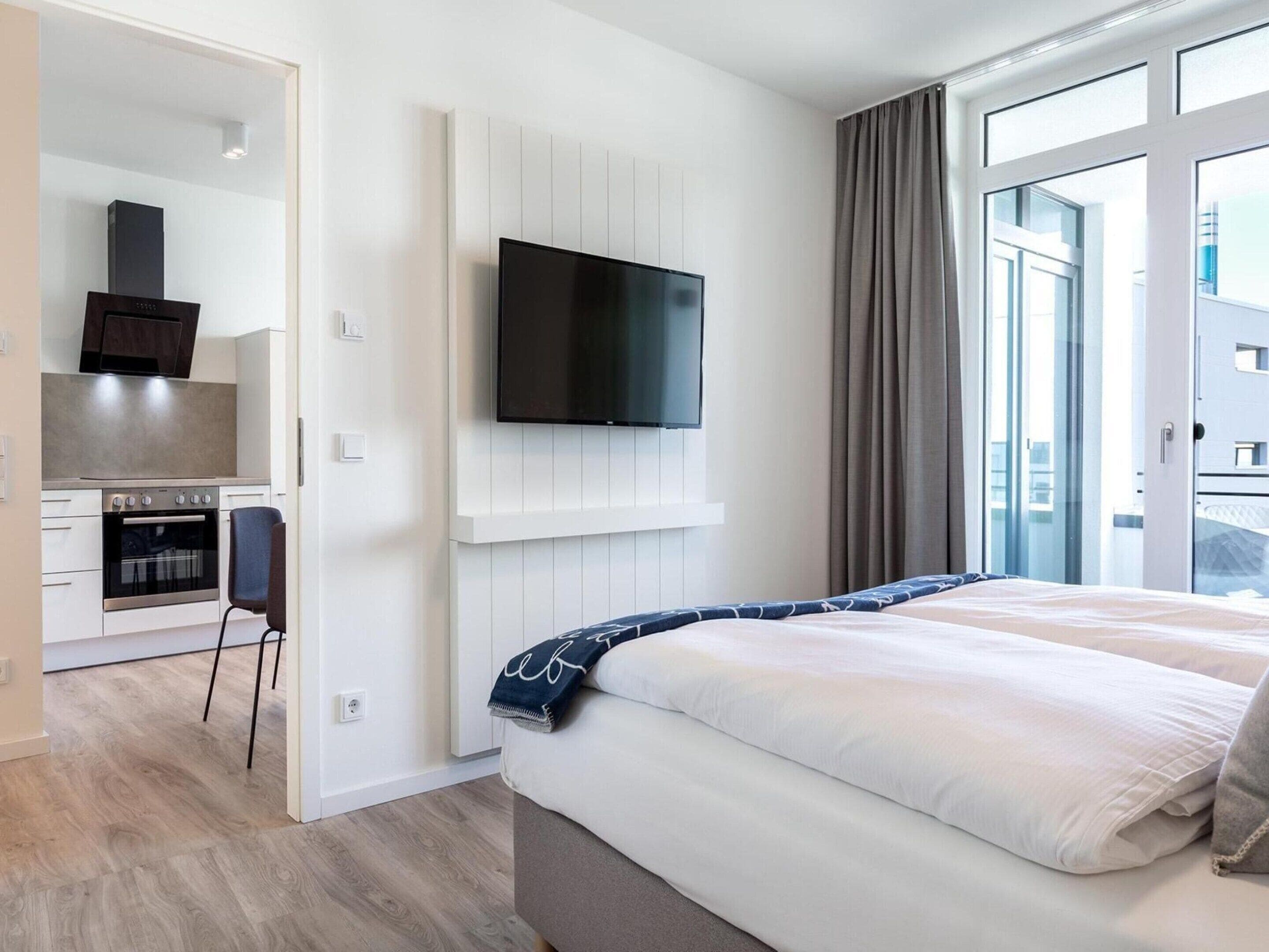Quarto de hotel moderno com uma cama, televisão na parede, cozinha compacta e uma janela grande.
