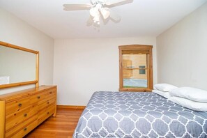 Um quarto com cama, cômoda de madeira, ventilador de teto e janela com persianas.