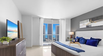 Quarto moderno com cama, televisão, escrivaninha e vista para edifícios altos.