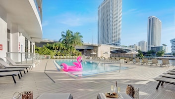 Área de piscina no terraço com espreguiçadeiras, um boi de flamingo flutuante e edifícios altos ao fundo.