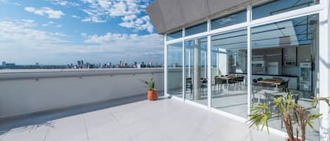 Um edifício moderno com amplas janelas de vidro, um terraço no telhado com área de refeições e vista para o horizonte da cidade.