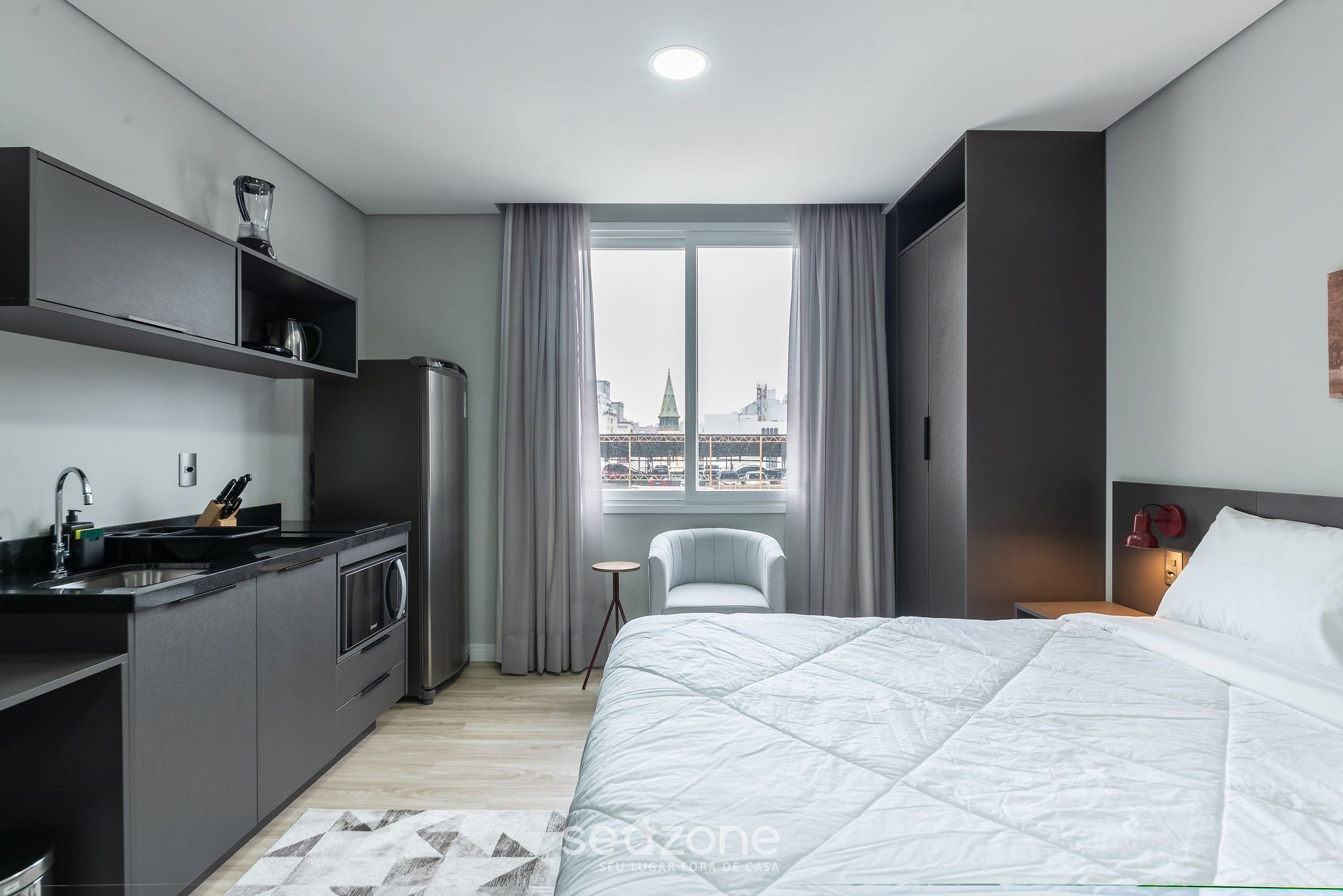Quarto de hotel moderno com cama, pequeno espaço de cozinha, janela com cortinas e vista para a cidade.