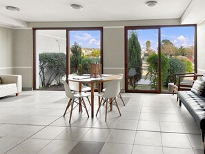 Uma sala de estar moderna com área de jantar, grandes portas de vidro deslizantes e vista para um jardim e prédios.