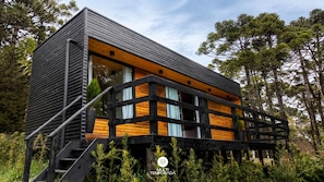 Uma cabana moderna com deck de madeira, grandes janelas e escada que leva à entrada.