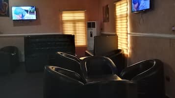 Quarto com televisão de tela plana na parede, teto com iluminação embutida e área de estar com cadeiras pretas.