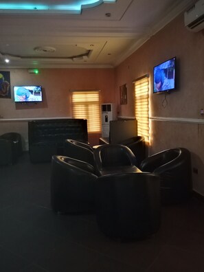 Quarto com televisão de tela plana na parede, teto com iluminação embutida e área de estar com cadeiras pretas.