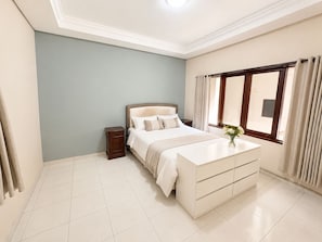 Quarto com cama, mesa de cabeceira, cômoda e janela com cortinas.