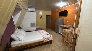 Quarto com cama, pequeno espaço de cozinha e porta que dá para outro cômodo.