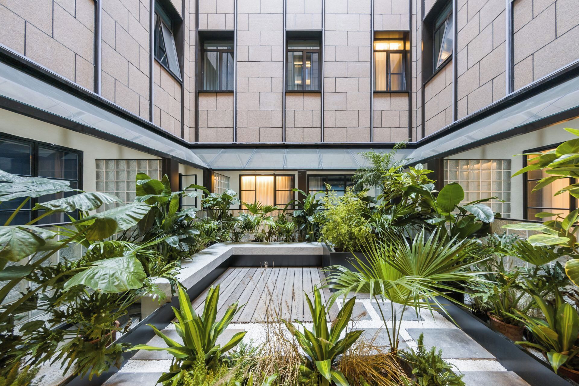 Um pátio com diversas plantas, um deck de madeira e fachada de um edifício moderno.