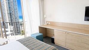 Quarto de hotel com cama, escrivaninha, cadeira, um banquinho e televisão na parede.